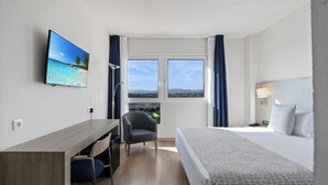 Quarto de hotel com cama, escrivaninha, cadeira, televisão e janela com vista para a praia e montanhas.
