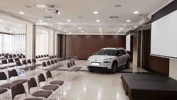 Uma sala de conferências com fileiras de cadeiras, um palco, um SUV branco e uma tela de projeção.