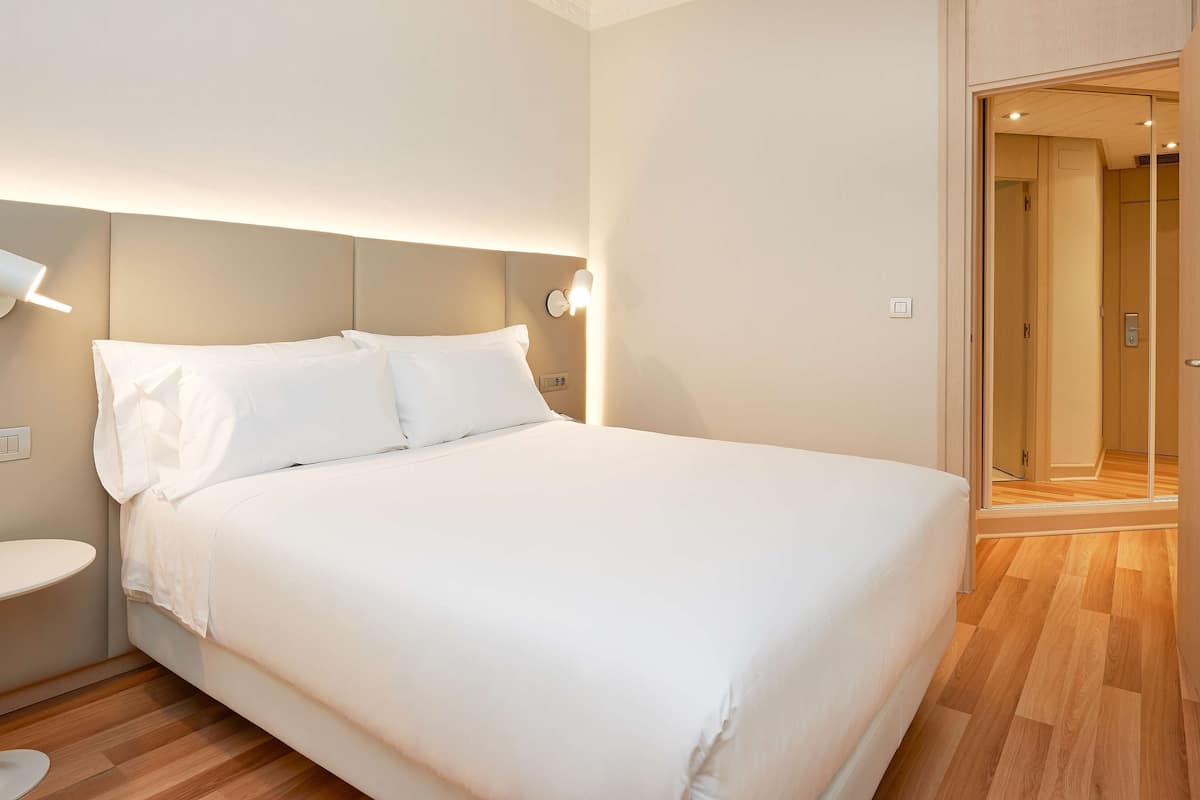 Cama bem arrumada com lençóis brancos em quarto moderno com piso de madeira e uma pequena mesa redonda.