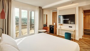 Quarto de hotel com uma cama grande, uma escrivaninha com TV, uma cadeira, um guarda-roupa e vista para a cidade pelas janelas.