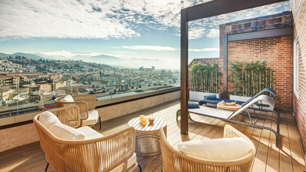 Varanda em prédio alto com móveis de vime, mesa redonda com bebidas e vista para a cidade.