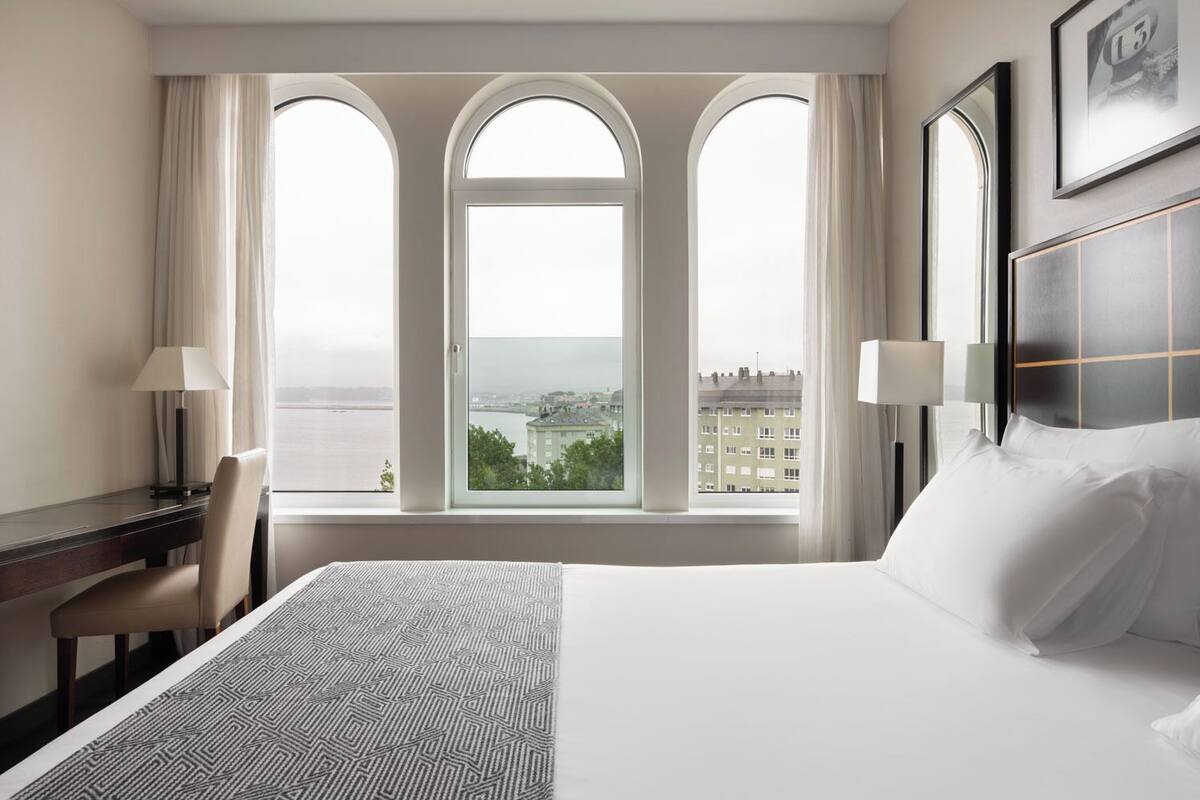 Quarto de hotel com uma cama grande, uma escrivaninha e uma cadeira. Três janelas oferecem vista para edifícios e para a água.