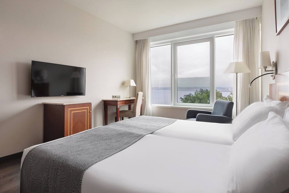 Quarto de hotel com uma cama grande, uma mesa com cadeira, uma televisão e uma janela com vista para a água.
