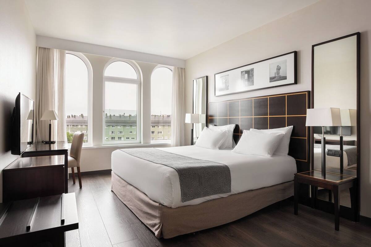 Quarto de hotel com uma cama grande, uma escrivaninha, uma cadeira, televisão e janela com vista para edifícios.