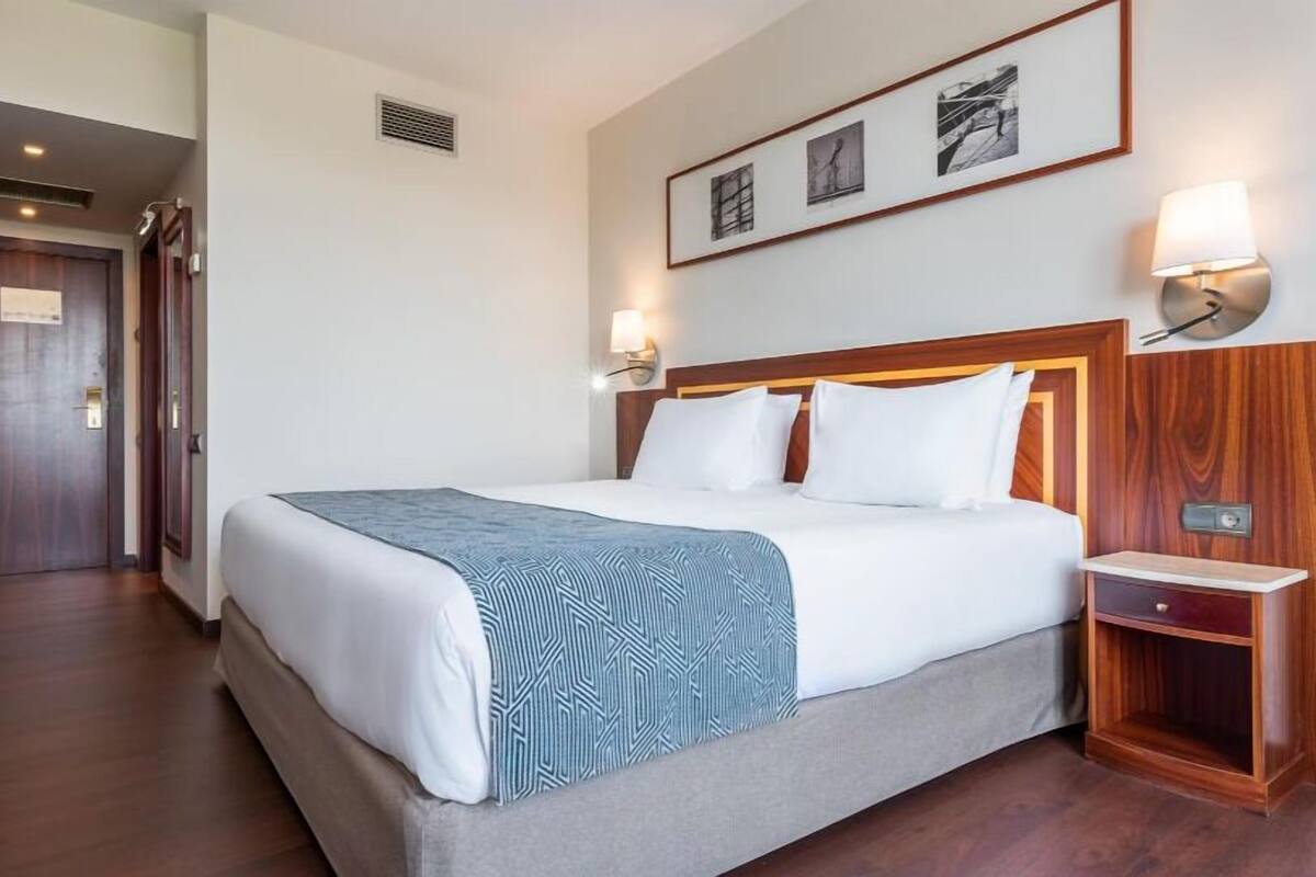 Quarto de hotel com uma cama grande, uma mesa de cabeceira e uma luminária fixada na parede.