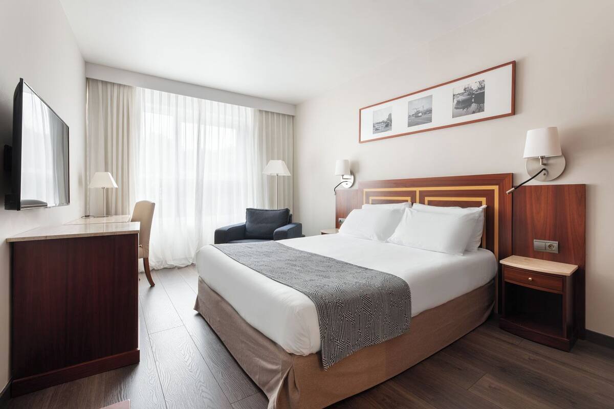 Quarto de hotel com cama, mesas de cabeceira, escrivaninha, cadeira, televisão e cortinas.