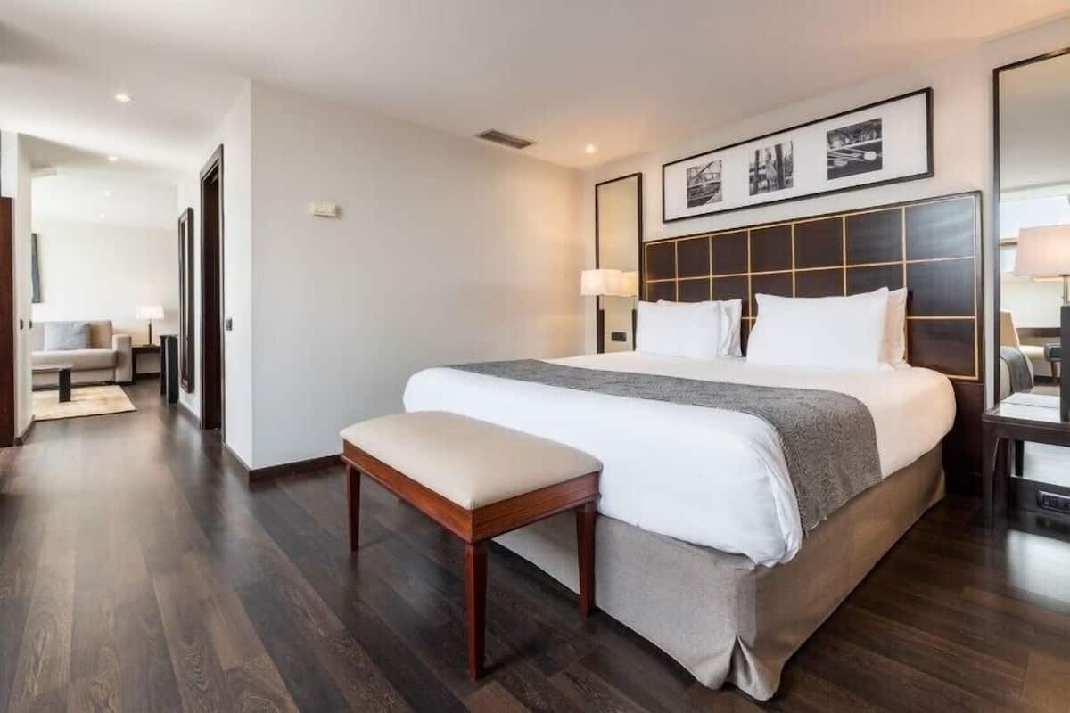 Quarto de hotel moderno com uma cama grande, uma mesa de cabeceira, um banco e uma cabeceira com um quadro emoldurado acima.