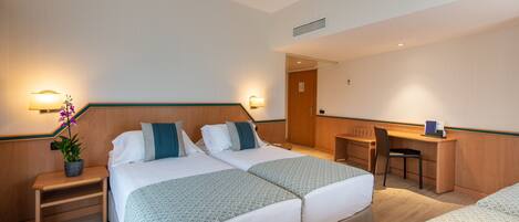 Quarto de hotel com duas camas, uma mesa de trabalho e uma cadeira. Há uma mesinha com uma luminária e uma planta.