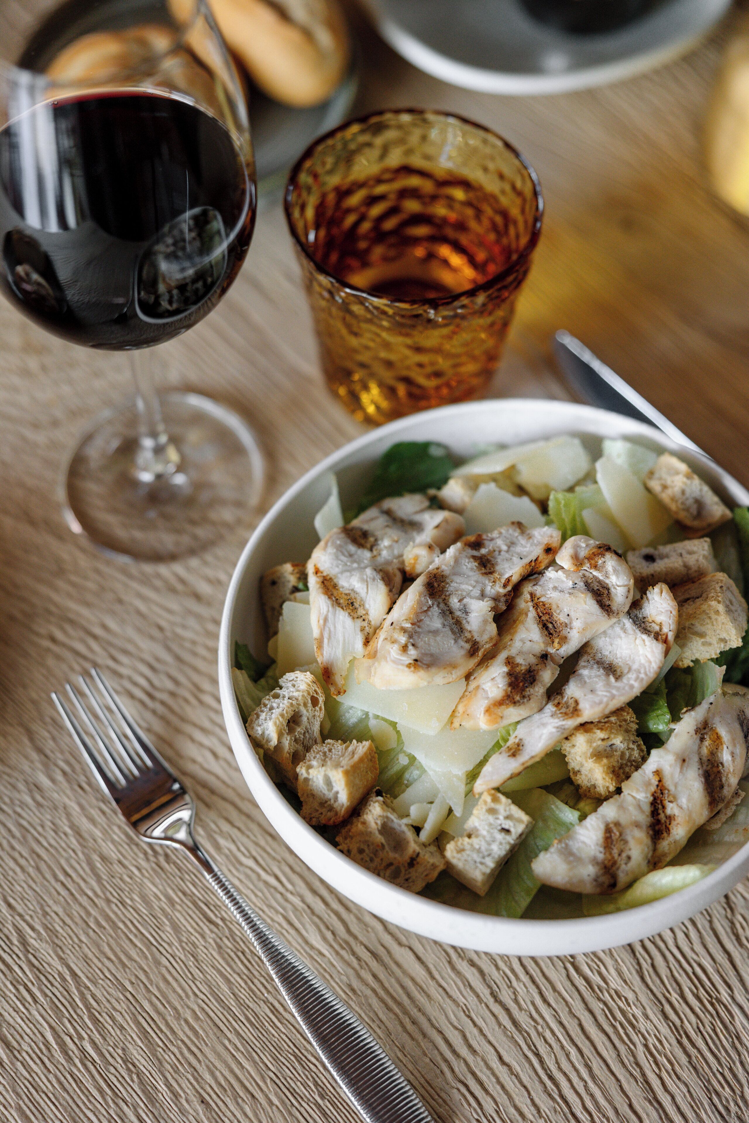 Uma salada com frango grelhado, um garfo e um copo de vinho tinto sobre uma mesa de madeira.