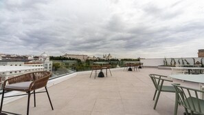Terraço no telhado com área de estar externa, guarda-corpo de vidro e vista para edifícios da cidade.