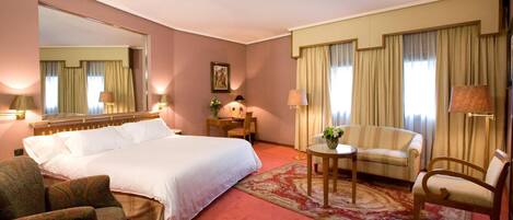 Quarto de hotel com uma cama grande, área de estar com sofá e poltronas, pequena mesa com um vaso, e um espelho amplo.