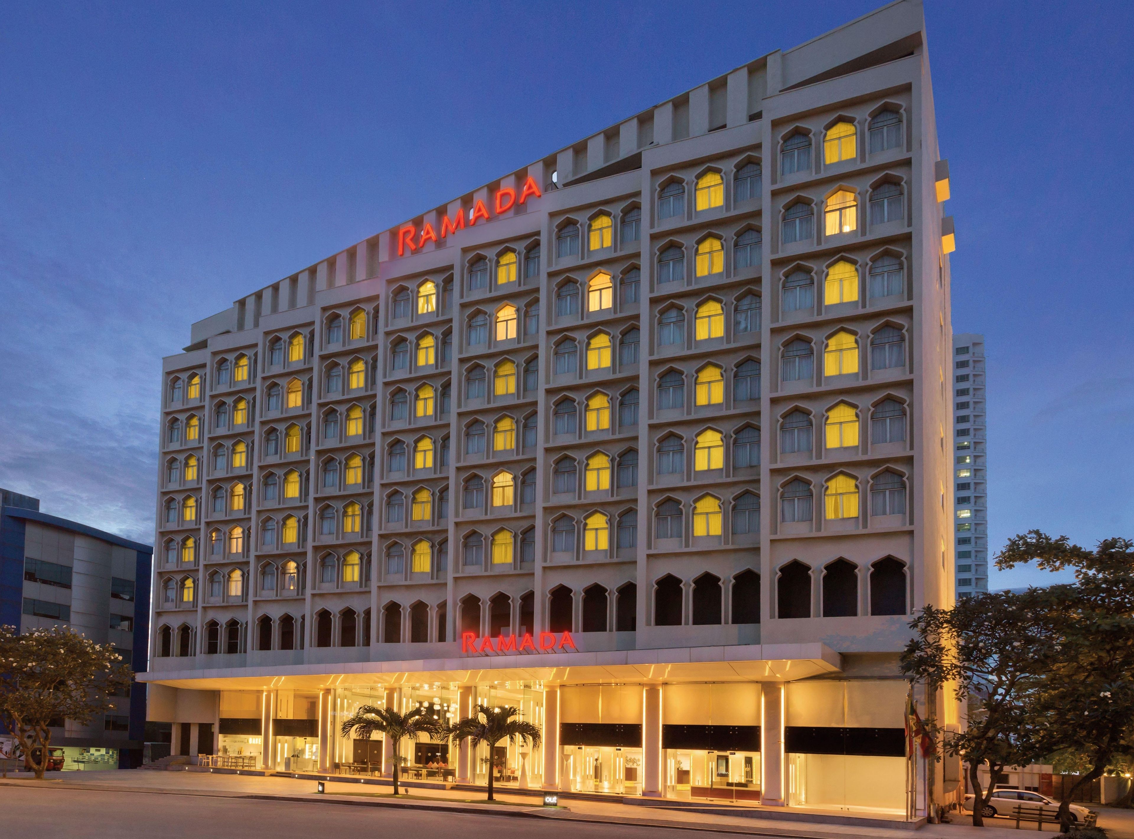 Um hotel de vários andares com uma placa grande da marca 'RAMADA', apresentando uma fachada simétrica com janelas uniformemente dispostas e uma entrada central.