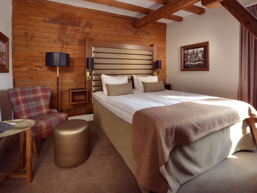 Quarto de hotel com teto de madeira, cama com roupa branca, poltrona xadrez, mesa redonda pequena com telefone e uma luminária.