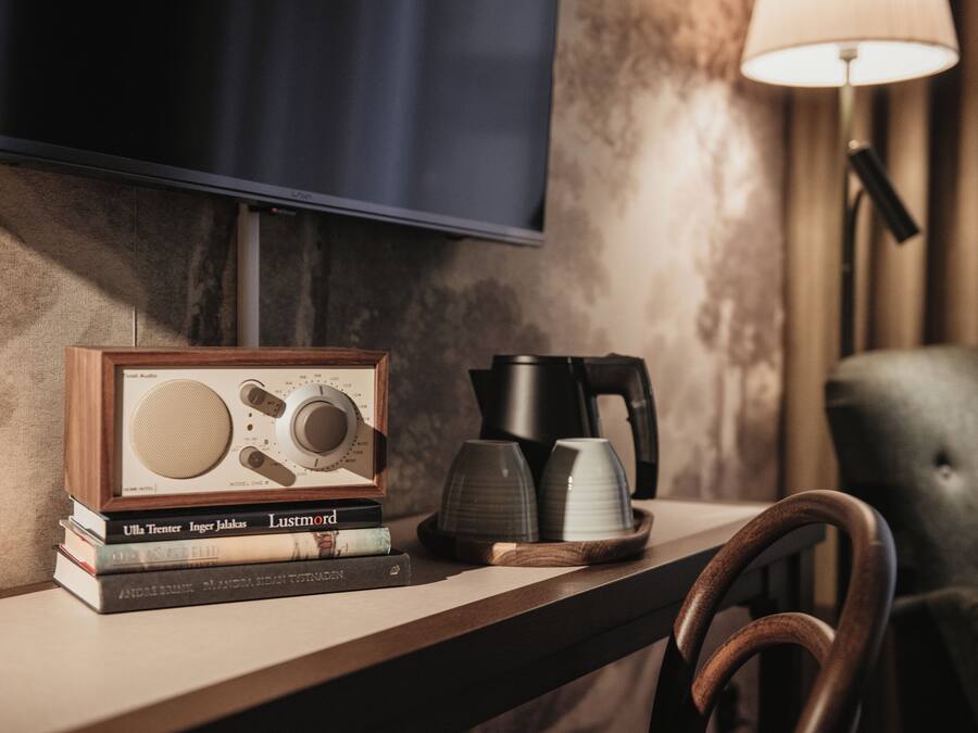 Um rádio de madeira, livros e uma cafeteira sobre uma mesa, com uma cadeira e um abajur ao fundo.