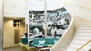 Um hall amplo com vista para a paisagem urbana através de uma grande janela, uma escadaria com corrimão dourado e uma área de estar com cadeiras verde-água e um tapete rosa.