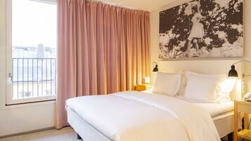 Quarto de hotel com uma cama grande, janela com cortinas, mesa de cabeceira com abajur e uma obra de arte pendurada na parede.
