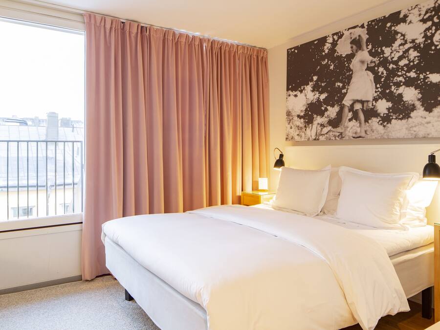 Quarto de hotel com uma cama grande, janela com cortinas, mesa de cabeceira com abajur e uma obra de arte pendurada na parede.
