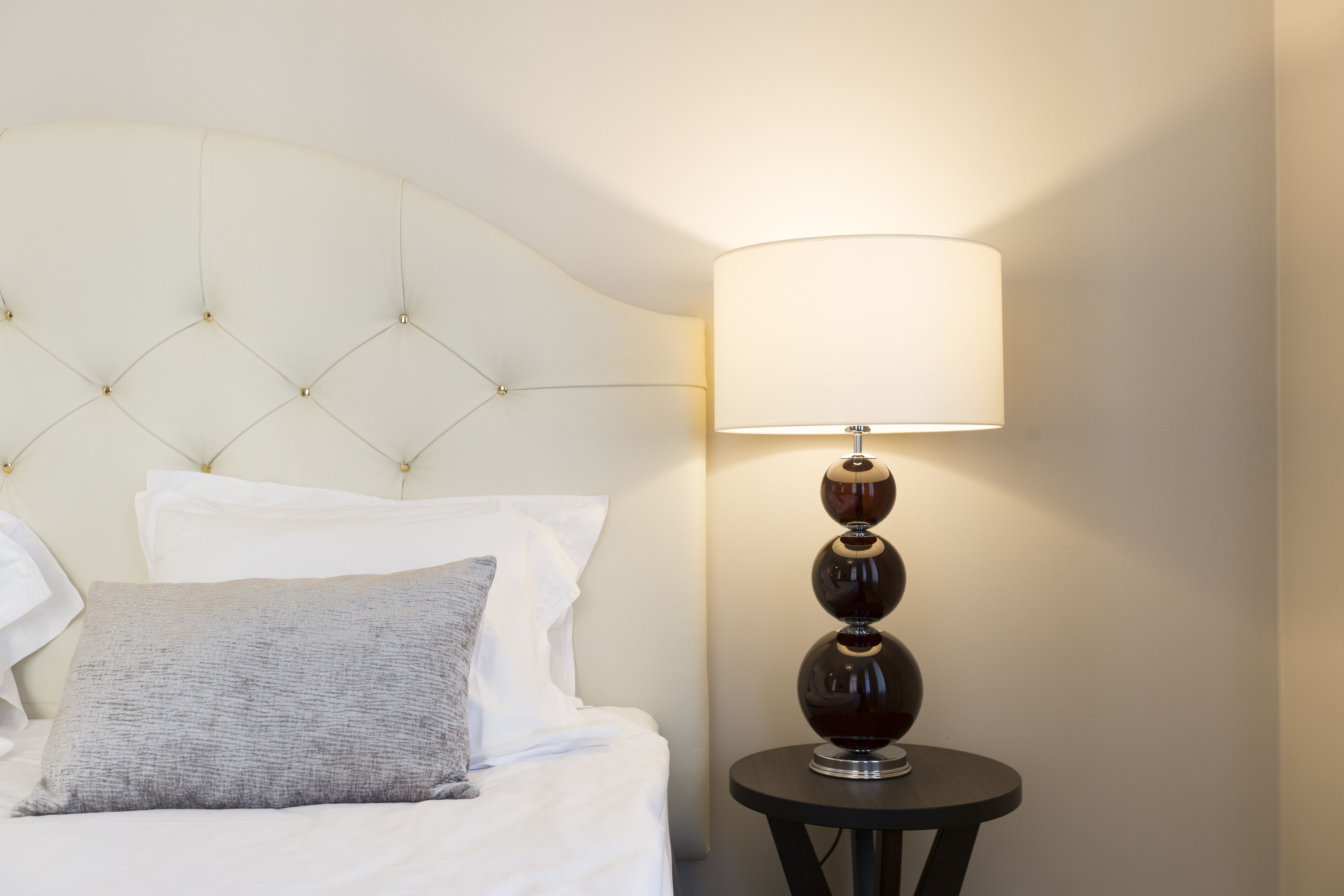 Uma cama bem arrumada com cabeceira estofada, uma luminária de cabeceira e uma pequena mesa redonda.