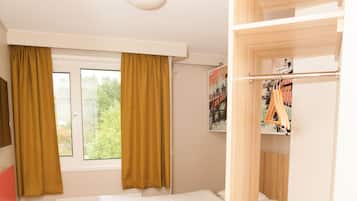 Quarto com cama, guarda-roupa, radiador, janela com cortinas e um pequeno banquinho.