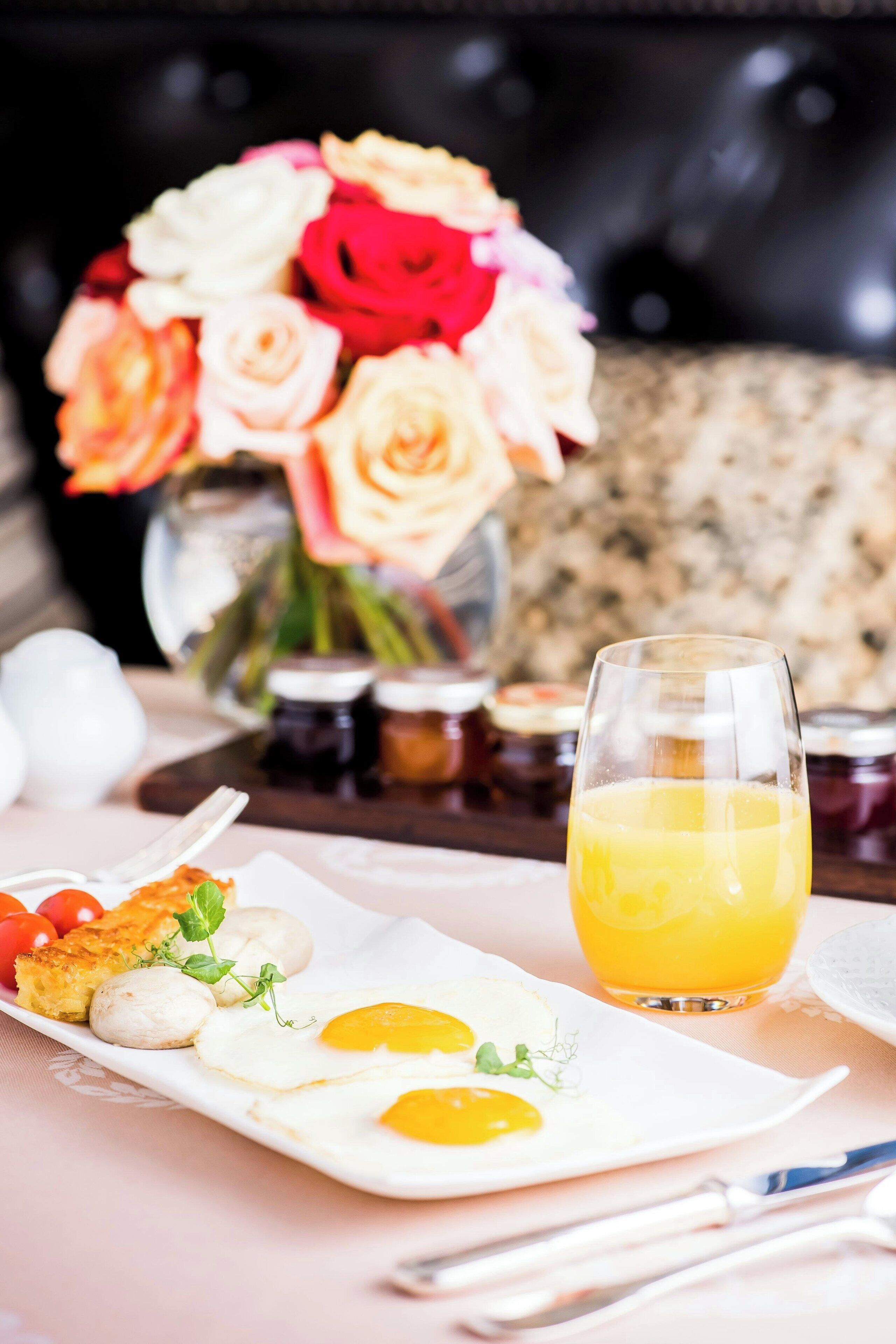 Mesa posta para o café da manhã com ovos, torradas e um copo de suco de laranja, com um vaso de flores ao fundo.
