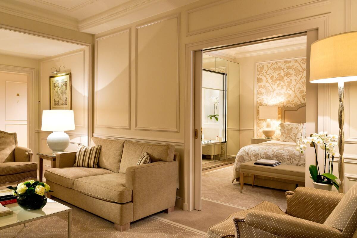 Quarto de hotel com sofá bege, uma mesinha com um vaso de flores, cama com edredom branco e uma luminária.