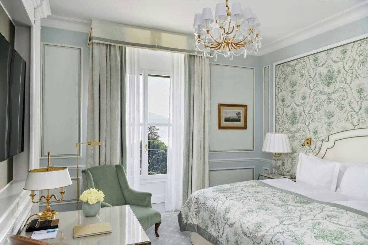 Quarto de hotel com cama, cadeira, escrivaninha com luminária, vaso com flores, quadro na parede e lustre.