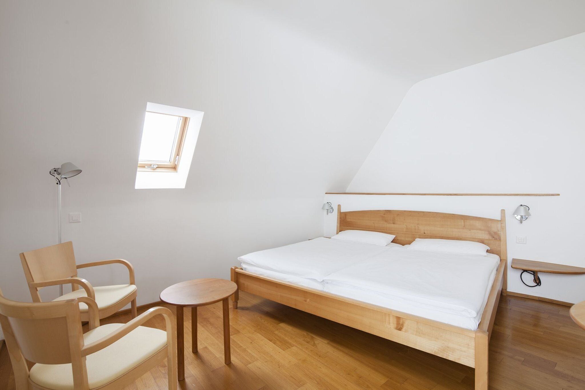 Quarto com cama de madeira, uma cadeira, uma mesinha, abajur e janela.