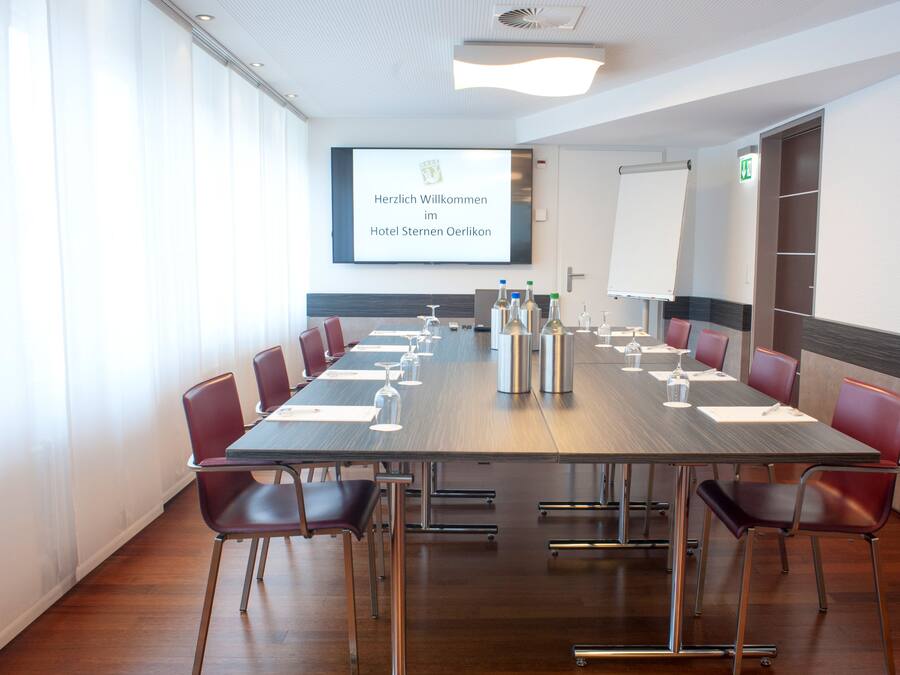 Uma sala de conferências com uma mesa comprida, cadeiras vermelhas e uma tela de projetor exibindo uma mensagem de boas-vindas.