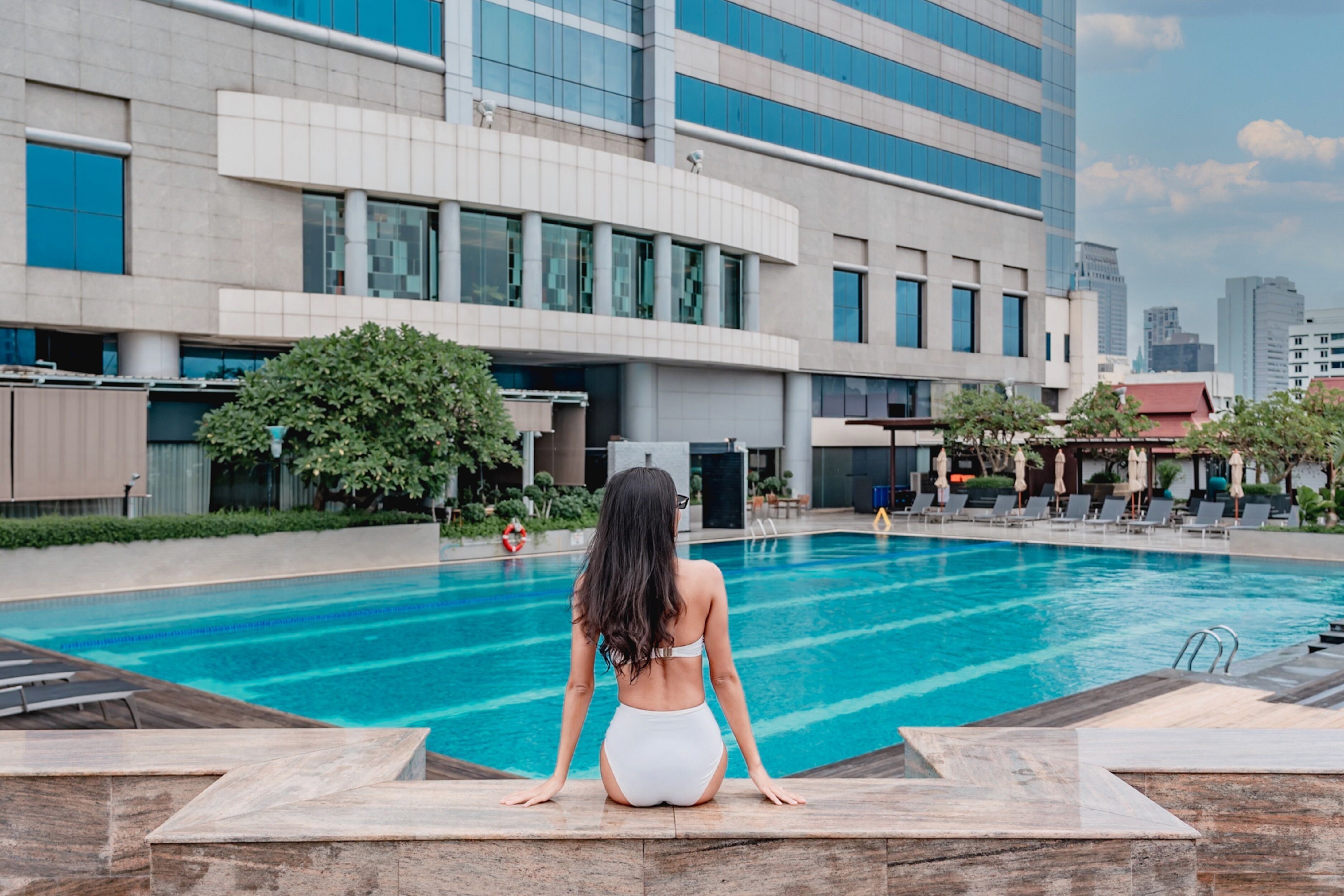 Uma mulher sentada à beira da piscina, com um edifício moderno ao fundo.