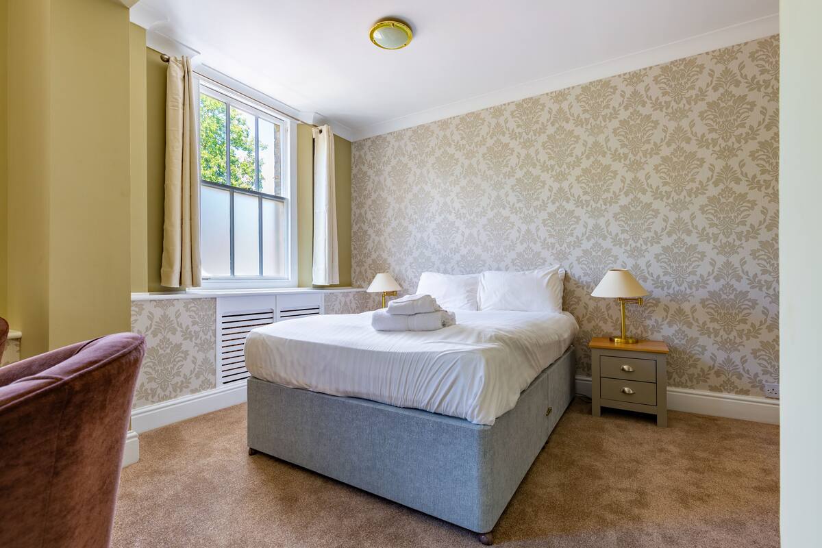 Quarto bem arrumado com cama, mesas de cabeceira, uma cadeira e janela com cortinas.