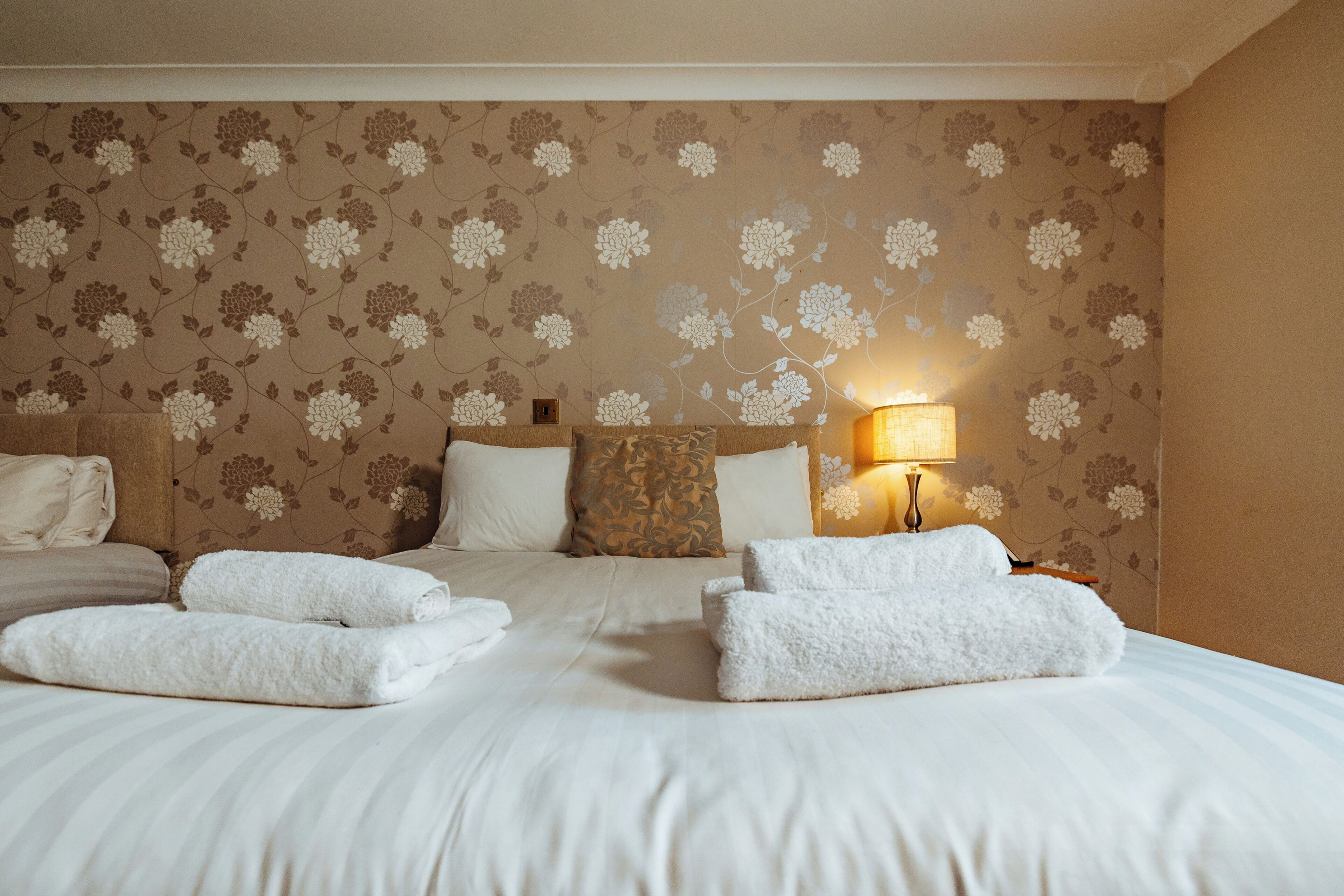 Quarto de hotel com papel de parede floral, duas camas com roupa de cama branca e uma luminária de cabeceira.