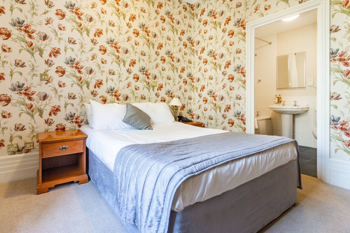 Quarto com papel de parede floral, uma cama com colcha cinza e branca, uma mesa de cabeceira com abajur, e um banheiro visível por uma porta aberta.