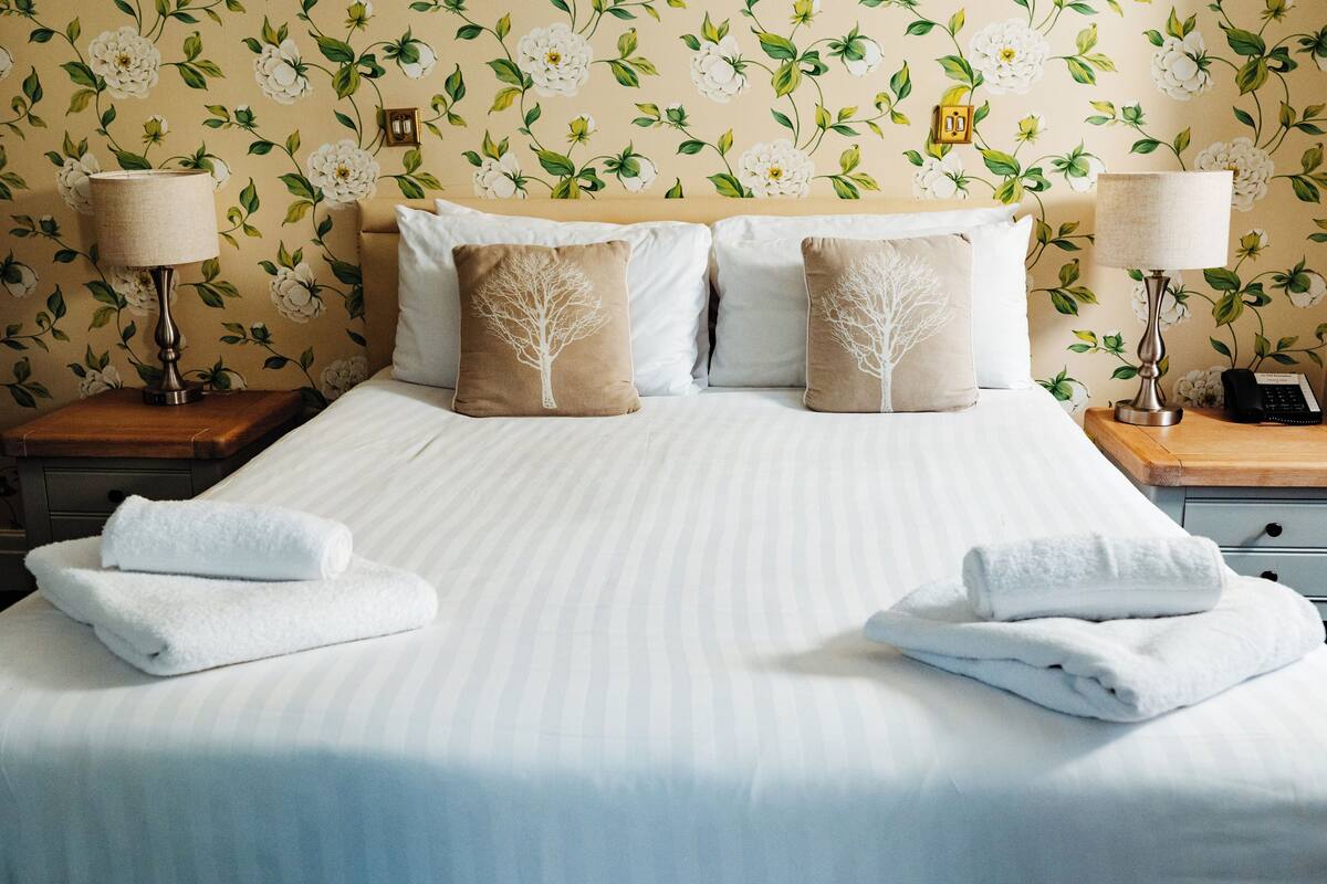 Uma cama bem arrumada com lençóis brancos, duas luminárias de cabeceira e papel de parede floral.