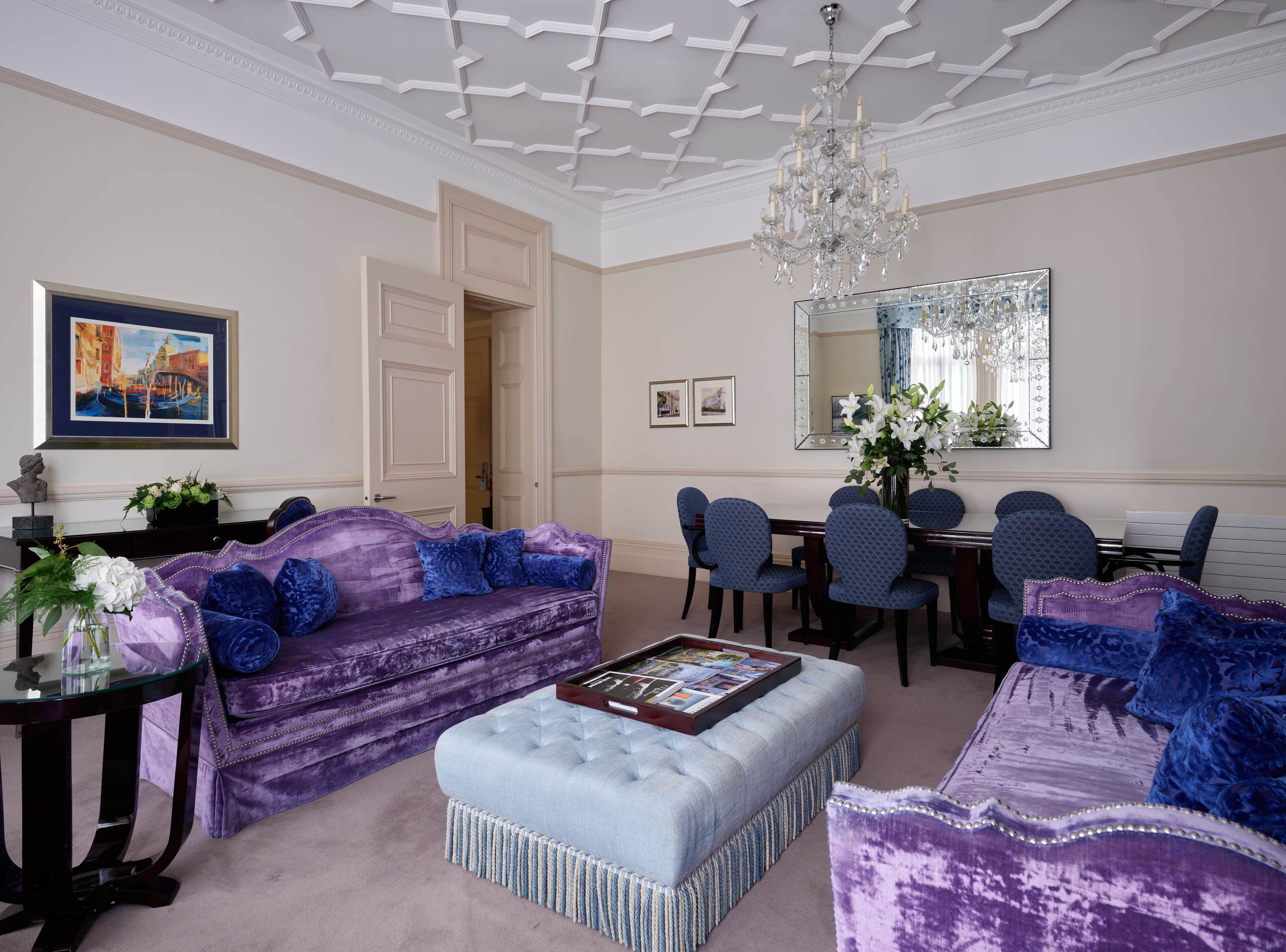 Uma sala de estar com um sofá roxo, um banquinho azul claro e uma área de jantar com cadeiras azuis e um lustre.