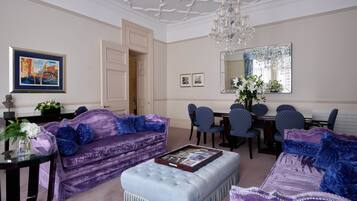 Uma sala de estar com um sofá roxo, um banquinho azul claro e uma área de jantar com cadeiras azuis e um lustre.