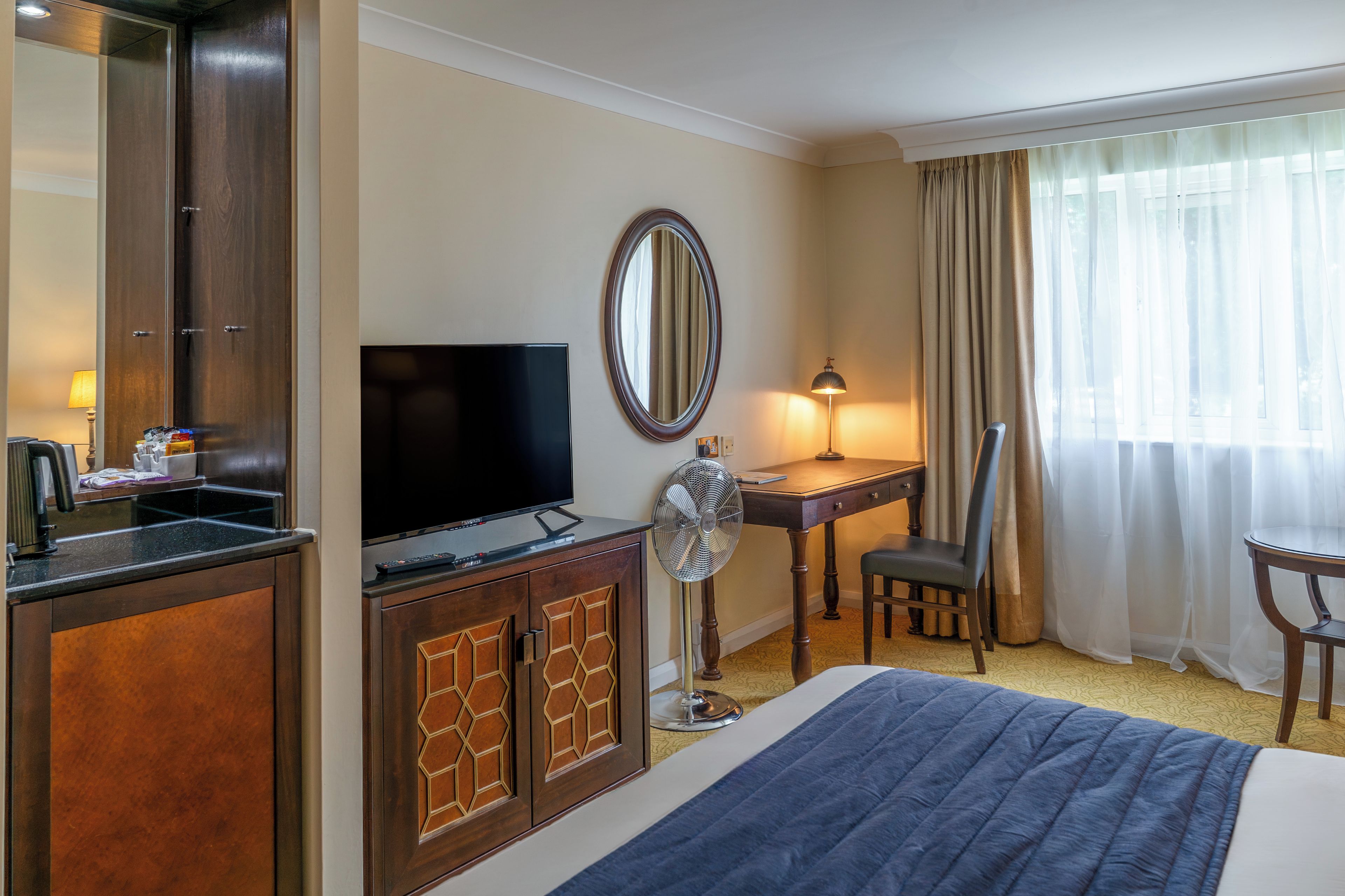 Quarto de hotel com cama, escrivaninha, televisão, espelho, luminária, cadeira e janela com cortinas.