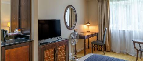 Quarto de hotel com cama, escrivaninha, televisão, espelho, luminária, cadeira e janela com cortinas.