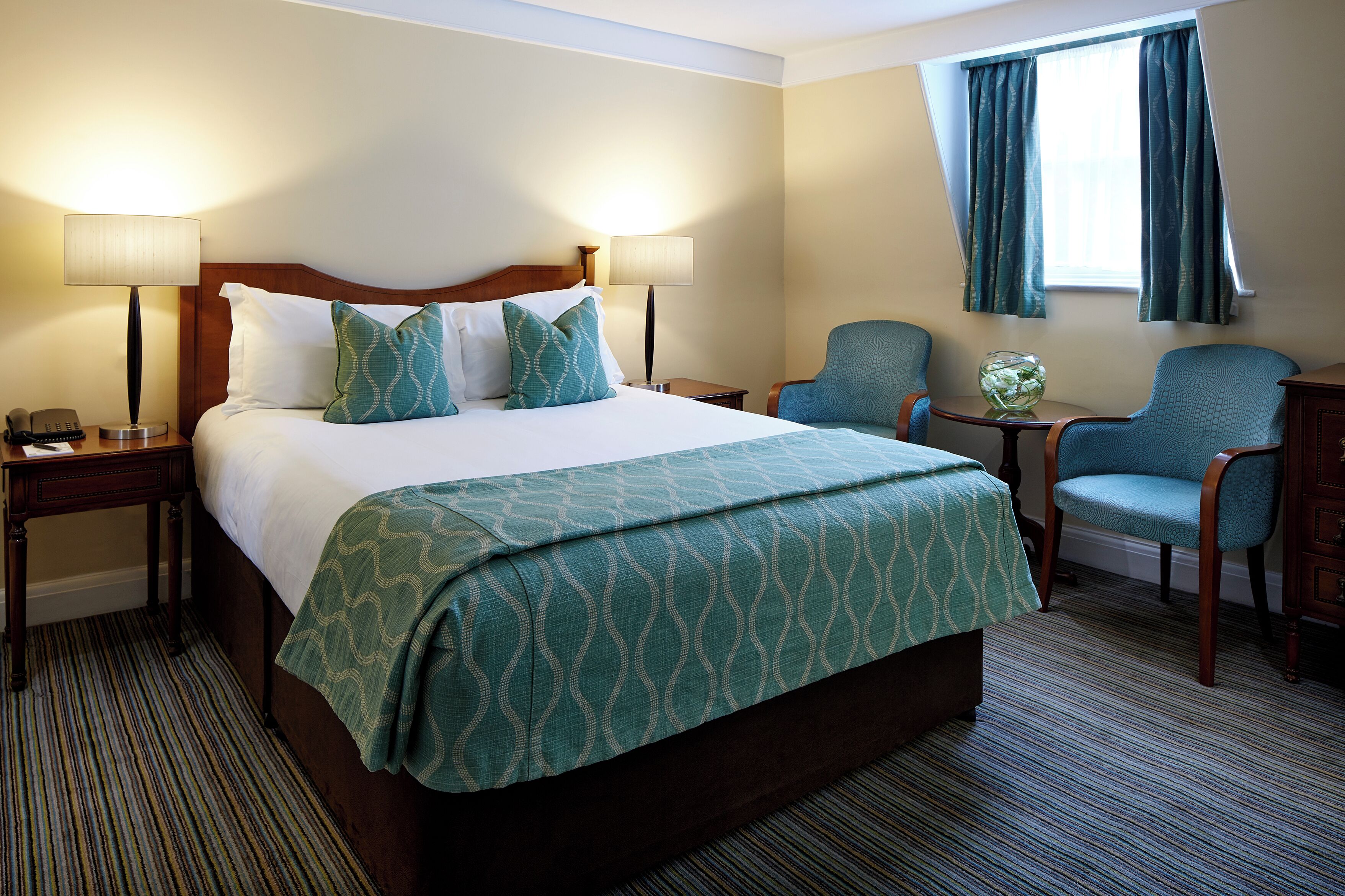 Quarto de hotel com uma cama grande, duas mesas de cabeceira, uma cadeira, uma mesinha e uma janela com cortinas.
