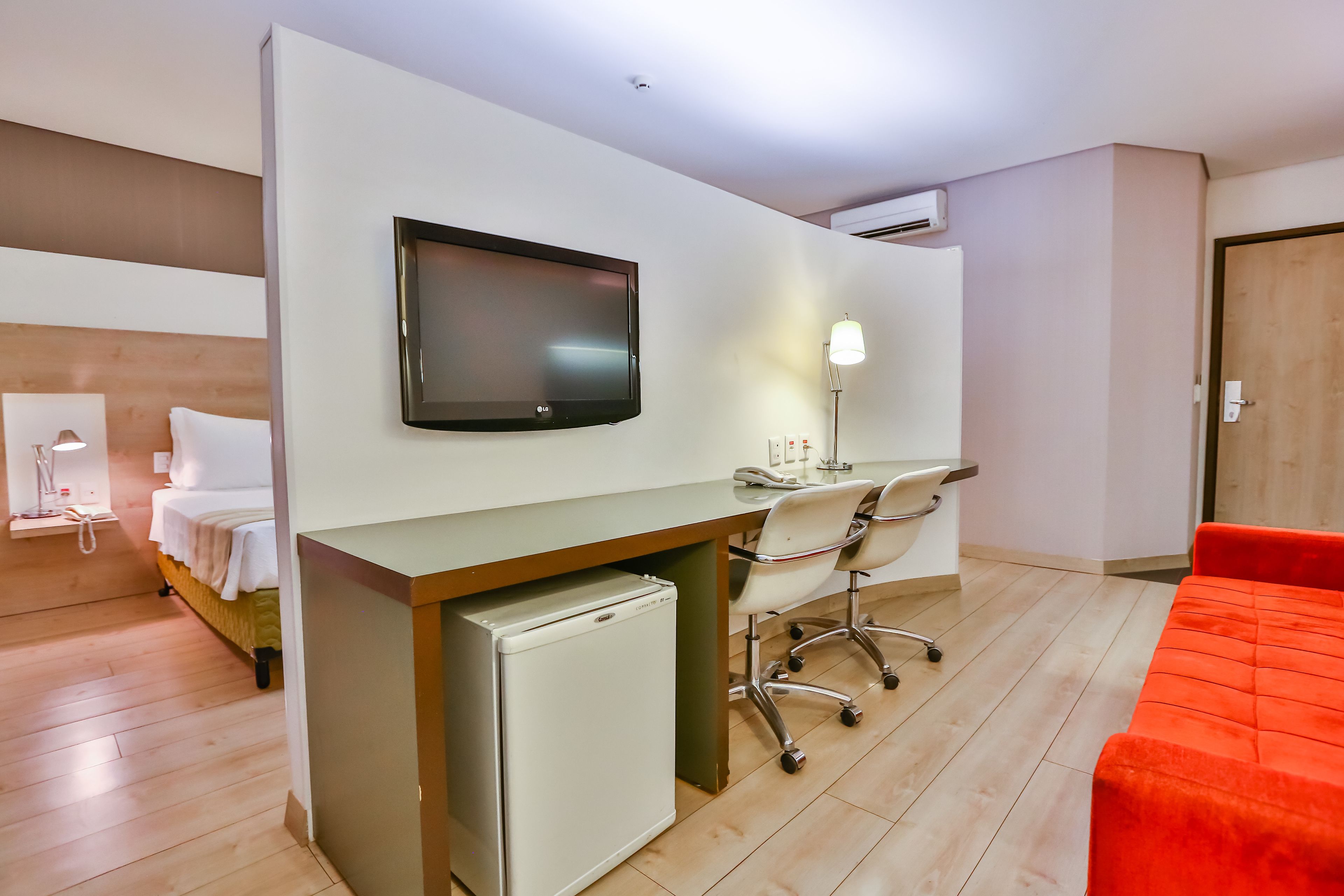 Quarto de hotel com cama, escrivaninha com cadeira, televisão, pequeno frigorífico e sofá vermelho.