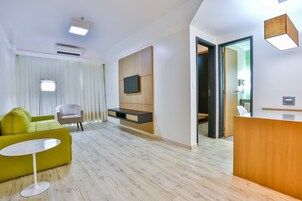 Quarto de hotel moderno com sofá verde, uma mesa redonda pequena, televisão de tela plana fixada em um painel de madeira e um balcão de recepção.