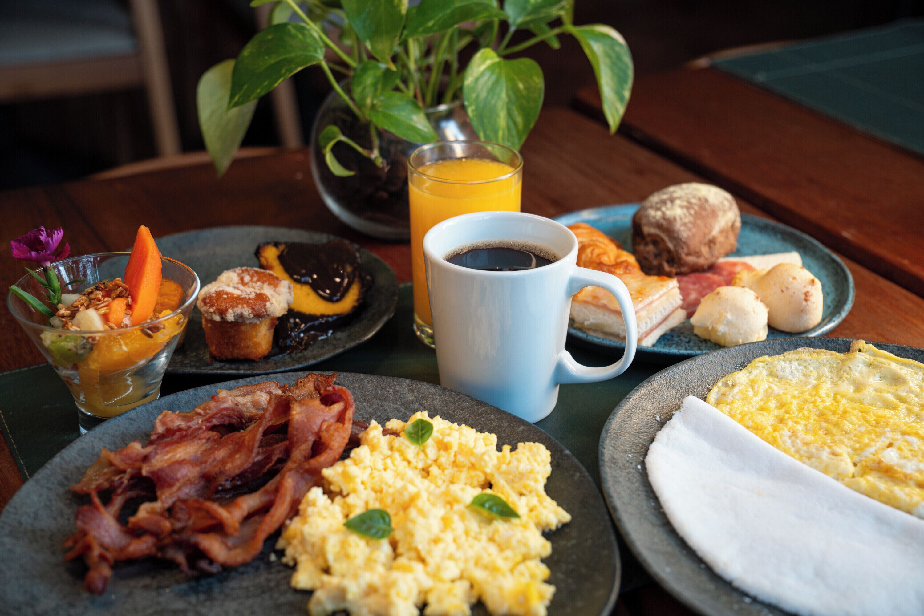 Uma mesa de café da manhã com diversos pratos, incluindo ovos, bacon, torradas e uma saladeira de frutas.
