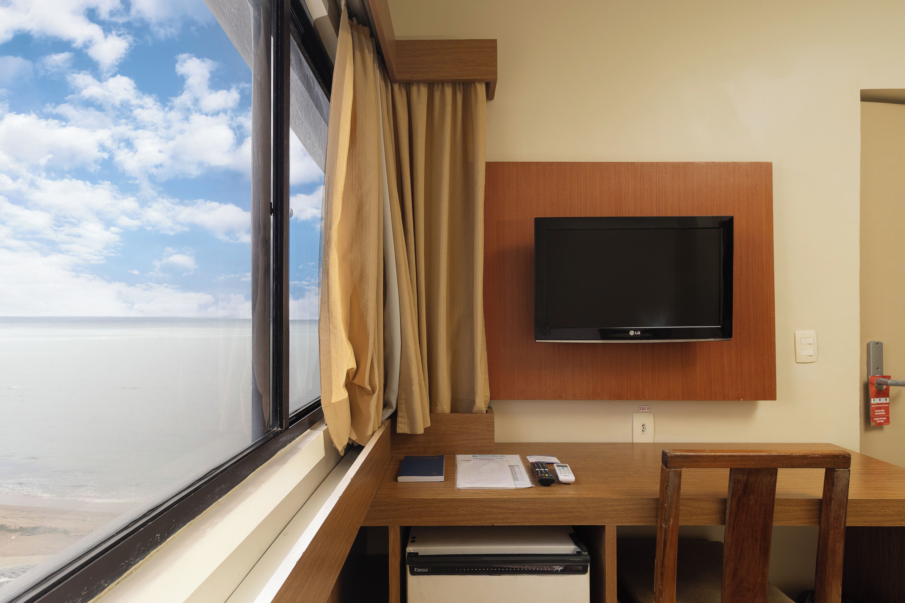 Quarto de hotel com uma janela grande, televisão de tela plana fixada na parede, escrivaninha de madeira com cadeira e uma mesa pequena com um controle remoto e um livro.