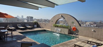 Piscina no terraço com vista para a cidade, cadeiras de descanso e uma pequena cascata.