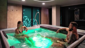 Piscina interior moderna com água cristalina, escada de metal e parede iluminada em verde.