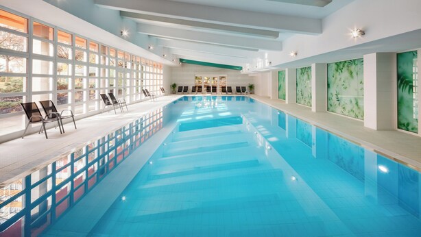 Uma piscina interior com paredes de vidro transparente, espreguiçadeiras e vista para o exterior.