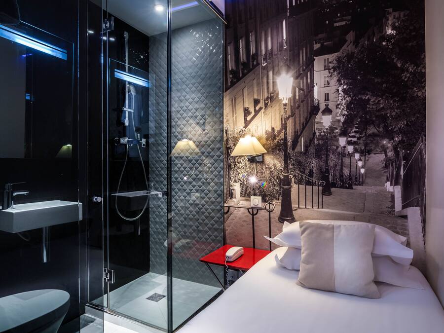 Quarto de hotel moderno com box de vidro, mesa de cabeceira com luminária e cama com almofadas.