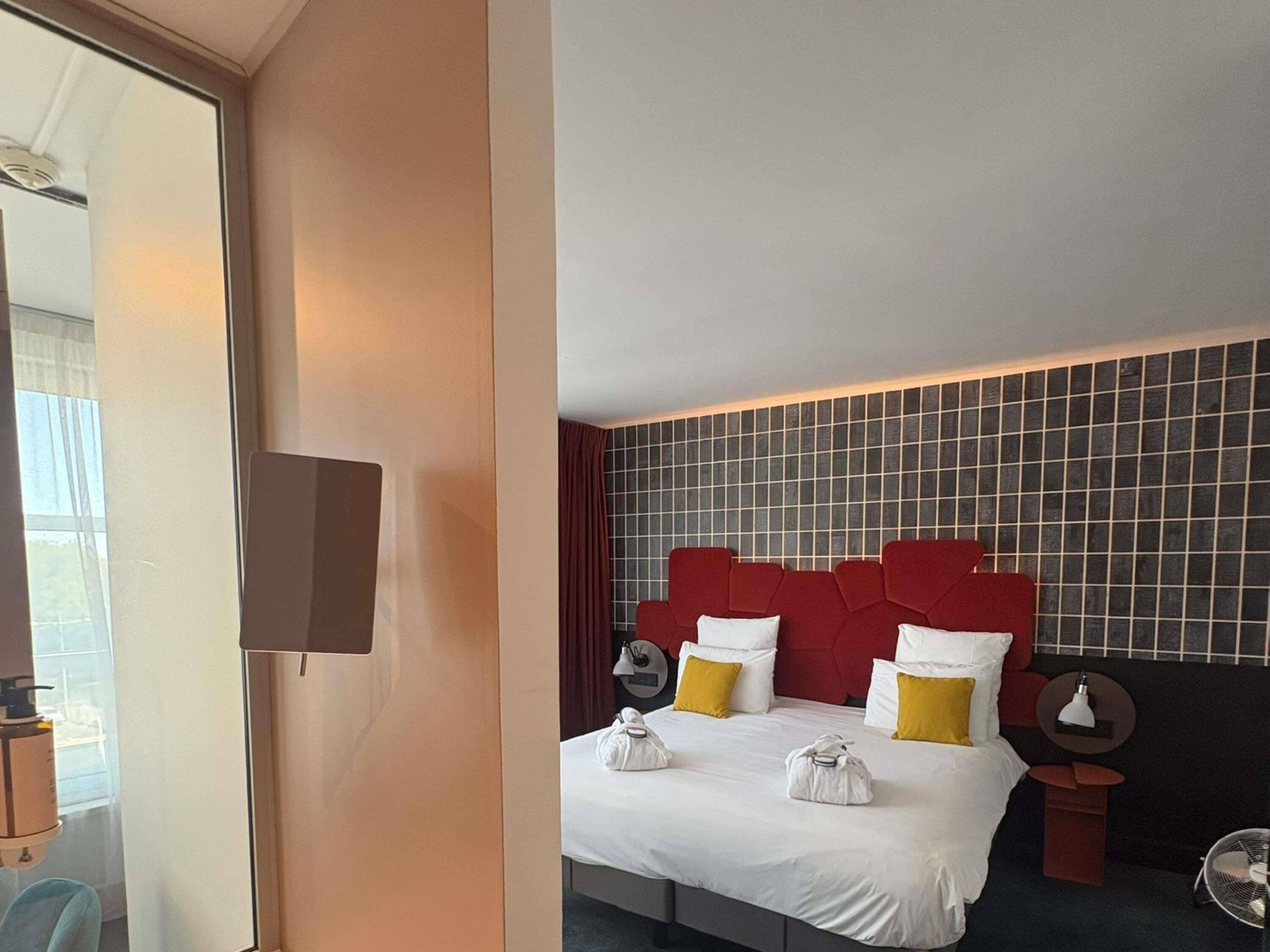 Quarto de hotel com cama, televisão na parede, mesa de cabeceira e janela.