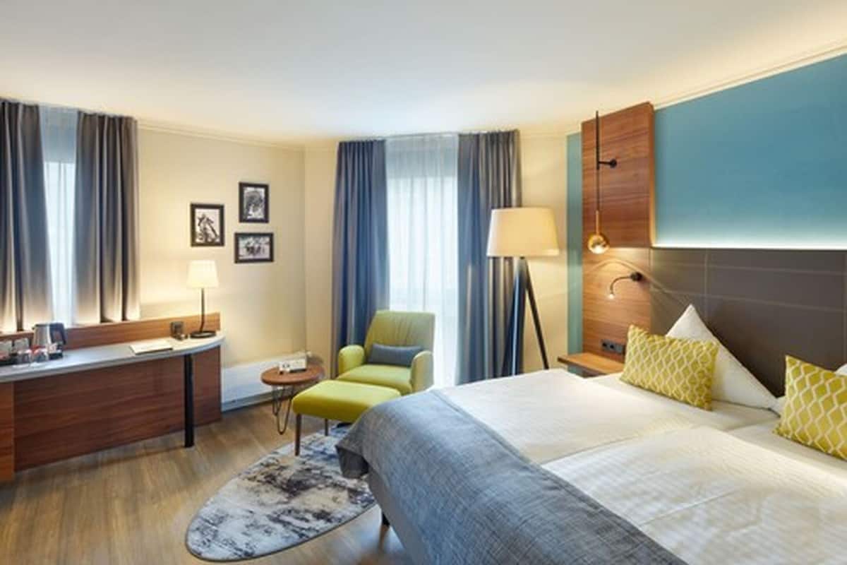 Quarto de hotel com cama, escrivaninha, cadeira, luminária e uma pequena mesa.