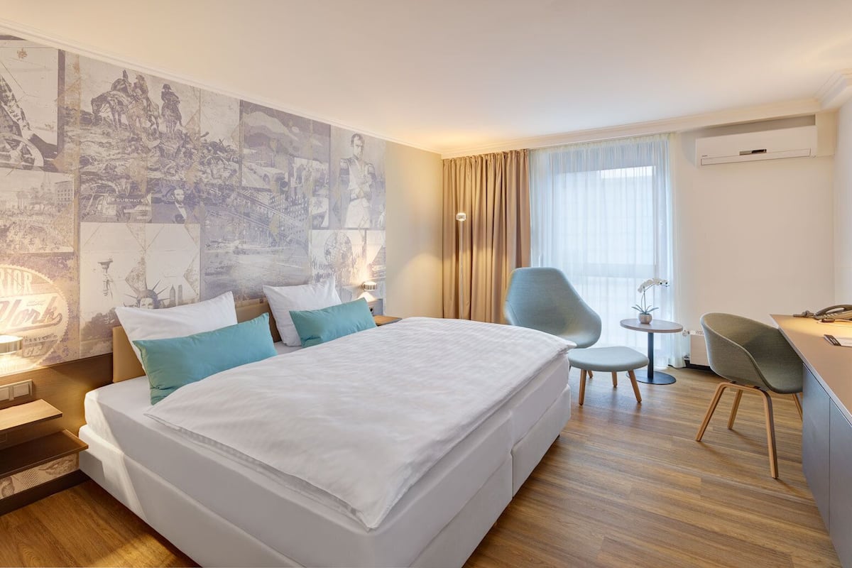 Quarto de hotel com uma cama grande, uma escrivaninha com cadeira, uma mesa pequena com cadeira e uma janela ampla com cortinas.