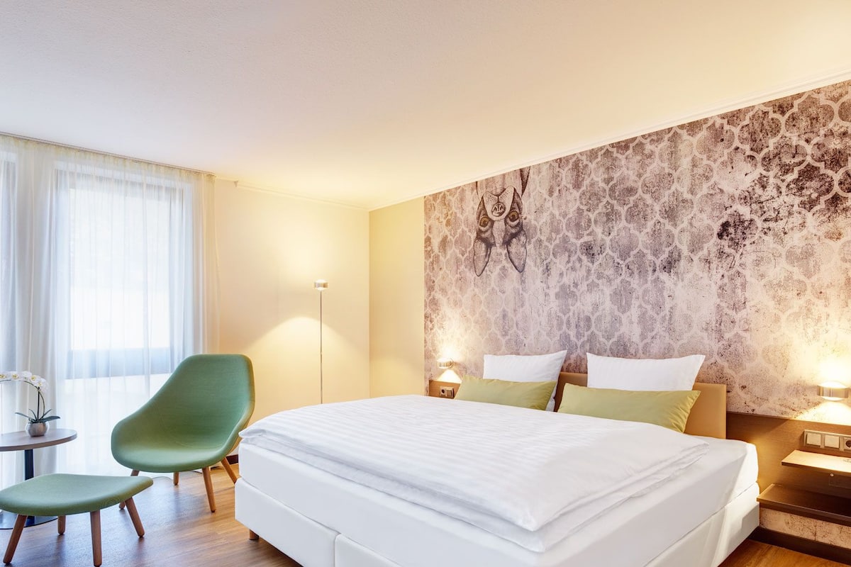 Quarto de hotel com uma cama grande, uma poltrona verde, uma mesinha com planta em vaso e uma parede com padrão texturizado.